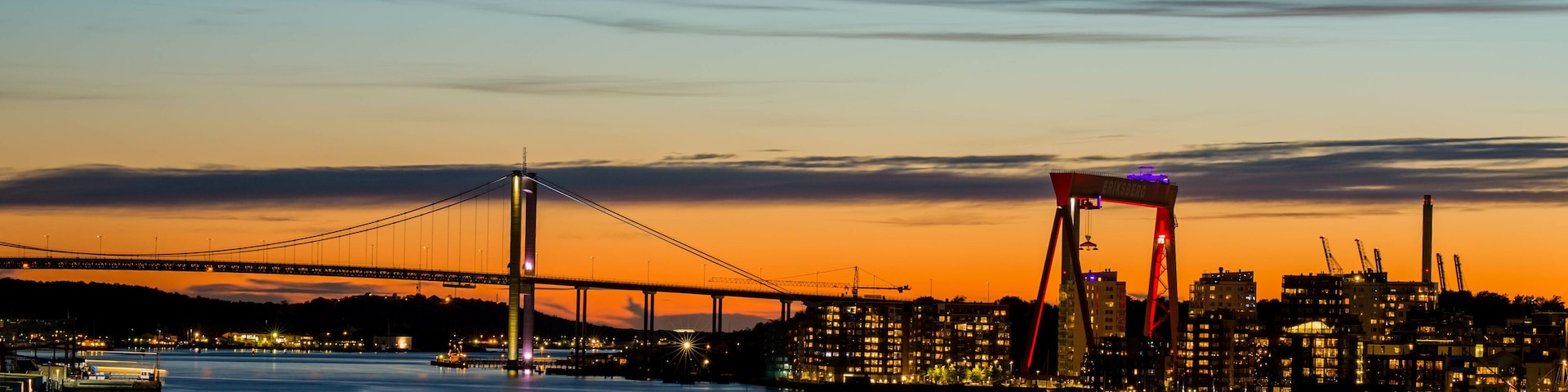 Paisagem urbana ao entardecer, com uma ponte imponente e uma torre de guindaste iluminada.