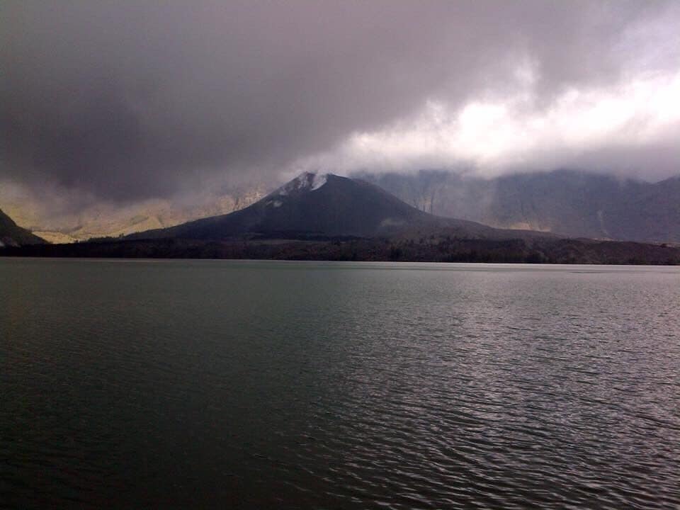Montanha parcialmente coberta por nuvens, um lago calmo e um céu nublado.