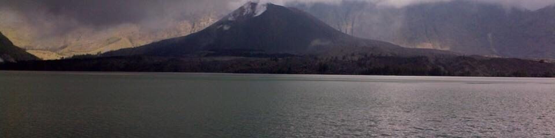 Montanha parcialmente coberta por nuvens, um lago calmo e um céu nublado.