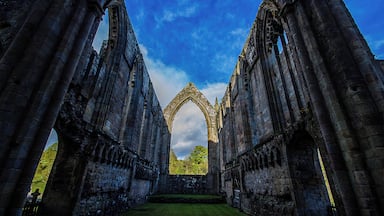 Um arco de pedra antiga com esculturas detalhadas, que leva a uma paisagem verde exuberante sob um céu azul límpido.