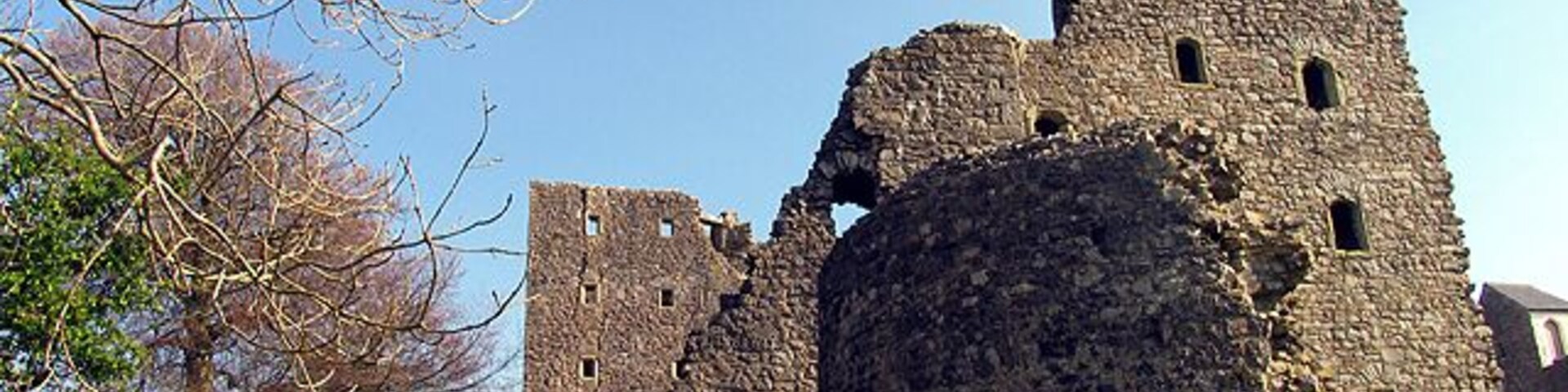 Ruínas de um grande castelo de pedra, com várias torres e um primeiro plano gramado.