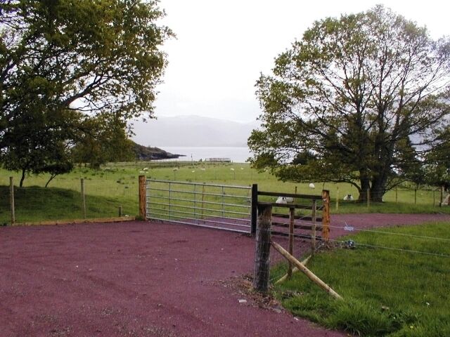 Paisagem rural com portão de metal, caminho vermelho e um campo gramado com árvores.