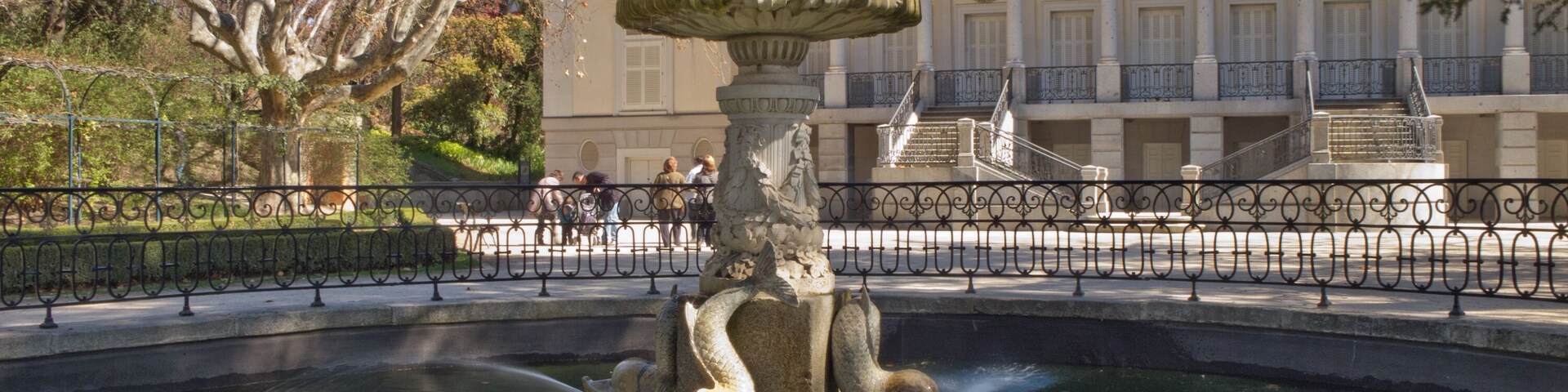 Uma fonte com estátua de peixe em um parque, com um edifício ao fundo.