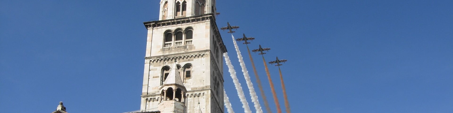 Igreja histórica com uma alta torre sineira e um grupo de aviões executando uma formação de voo com rastros de fumaça colorida.