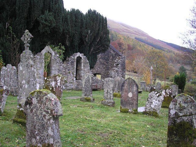 Um cemitério com lápides de pedra antigas e uma igreja em ruínas ao fundo.