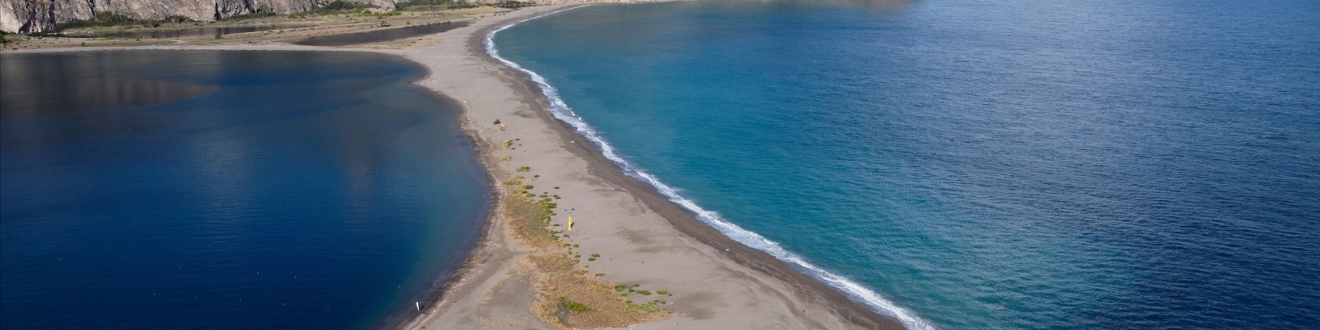 Paisagem costeira com uma praia arenosa, mar azul cristalino e uma colina rochosa com uma construção no topo.