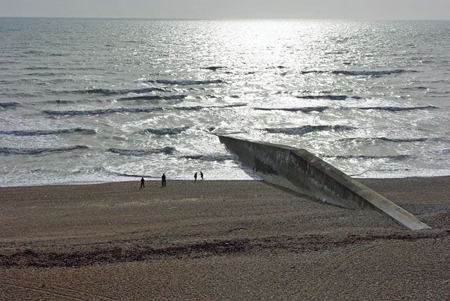 Uma praia com ondas, uma barreira de concreto e algumas pessoas.