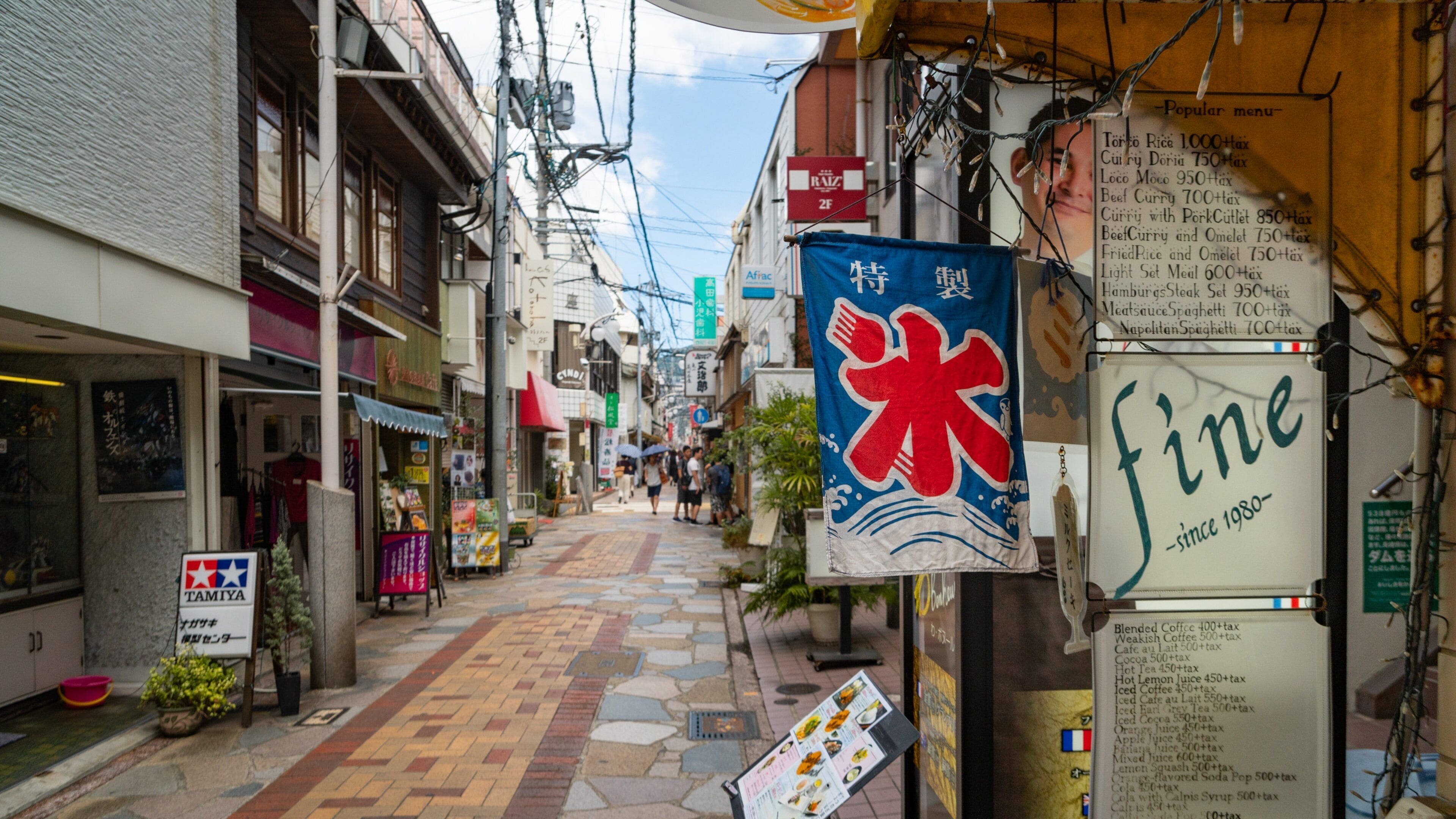 Uma rua estreita, flanqueada por lojas e restaurantes, com uma placa azul e vermelha com caracteres japoneses em destaque.