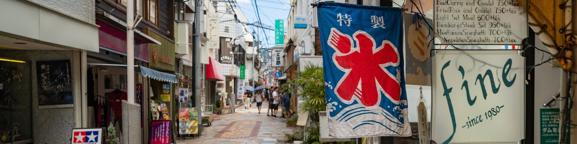 Uma rua estreita, flanqueada por lojas e restaurantes, com uma placa azul e vermelha com caracteres japoneses em destaque.