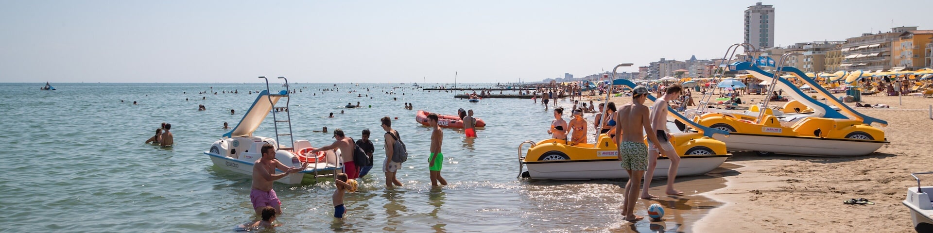 Uma praia cheia de gente nadando e permanecendo na água, com vários barcos amarelos e brancos estacionados na areia.
