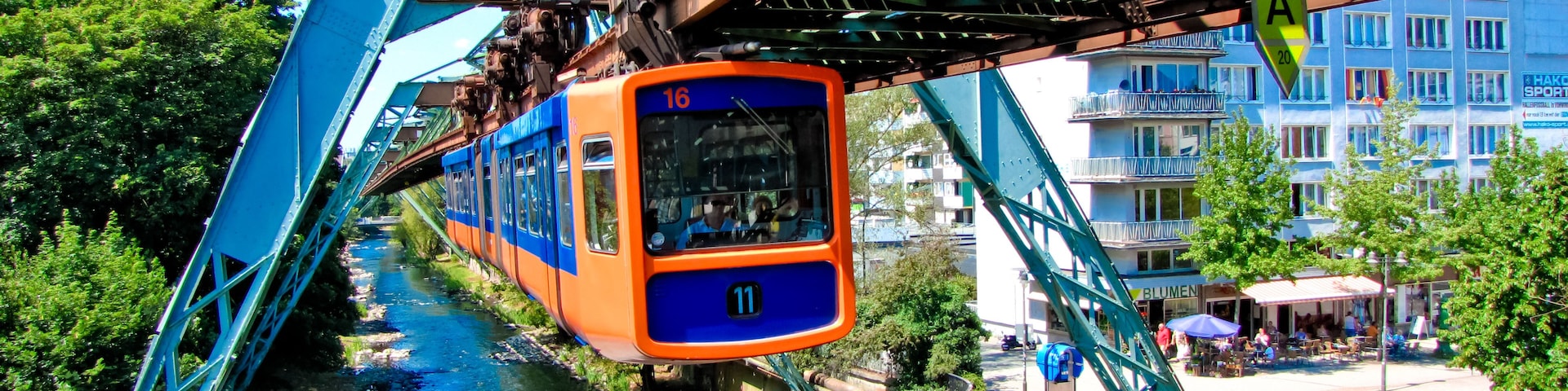 Um teleférico azul e laranja atravessa uma ponte sobre um rio, com edifícios e árvores ao fundo.