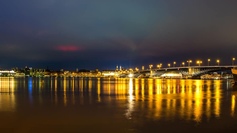 Cena noturna de um rio com uma ponte, edifícios iluminados e um barco atracado à direita.