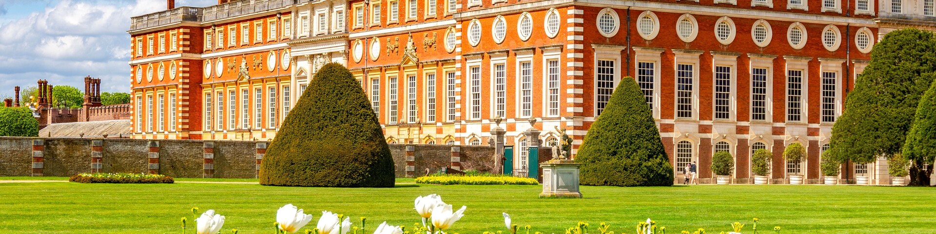 Um edifício grande de tijolos vermelhos com molduras de janela brancas, cercado por um gramado bem cuidado com tulipas brancas e roxas.
