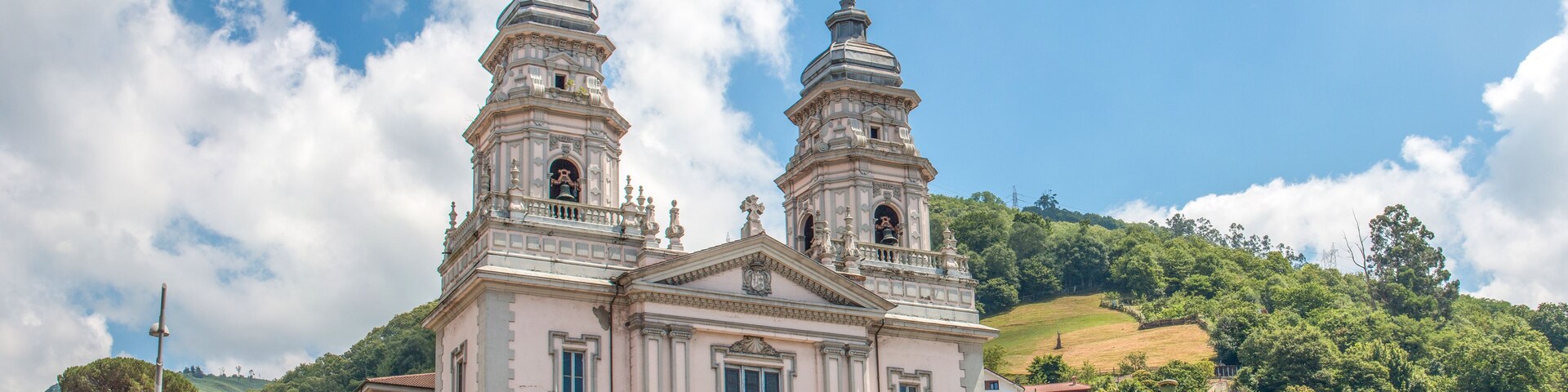 Igreja histórica com duas torres sineiras e uma cúpula, rodeada por colinas verdes e edifícios residenciais.