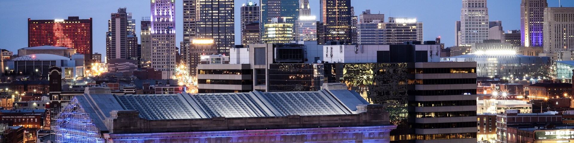 Silhueta urbana ao entardecer, com edifícios iluminados e um edifício histórico imponente com janelas em arco.