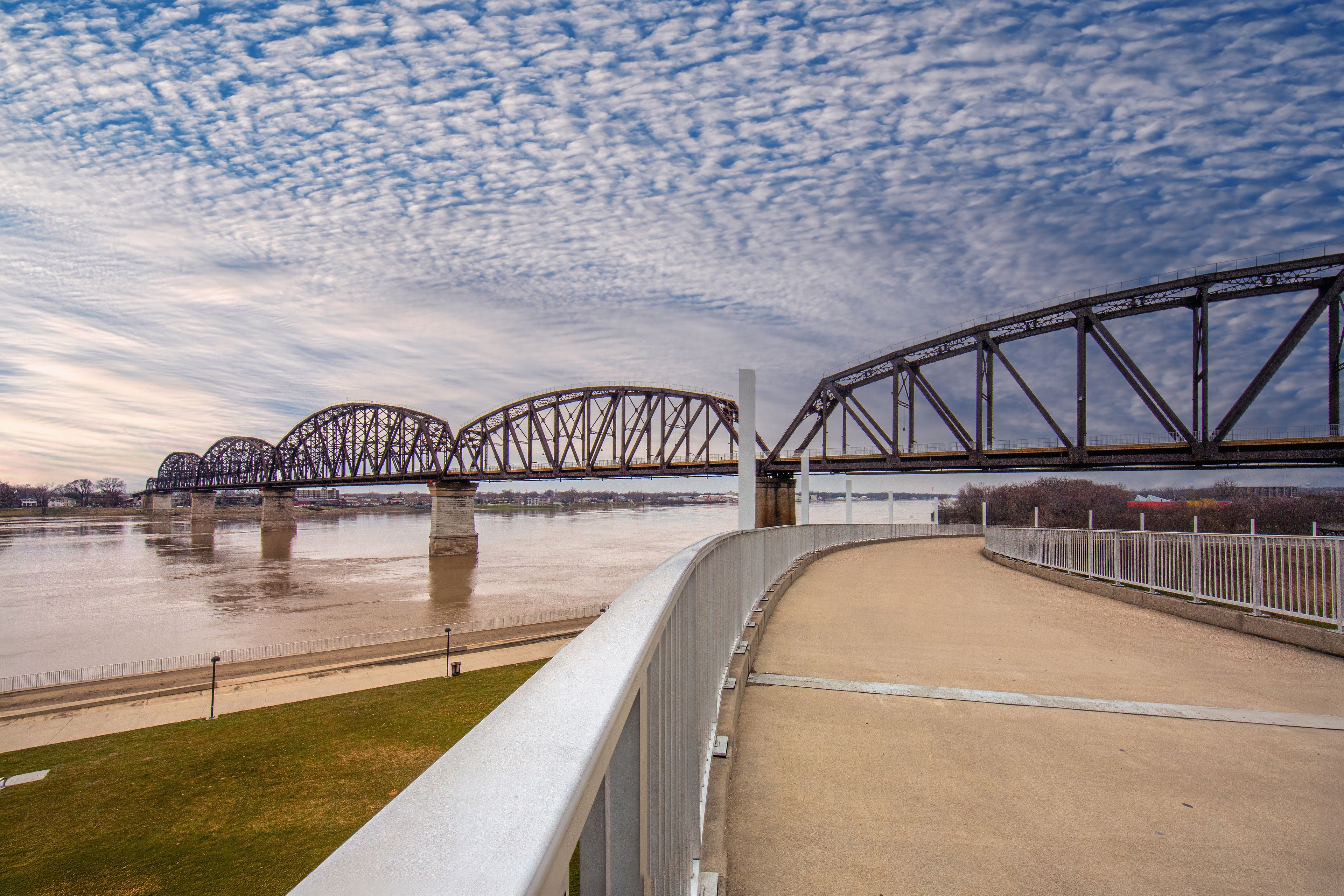 Uma ponte de aço com várias arcos sobre um rio, uma plataforma de observação de concreto e um céu nublado.