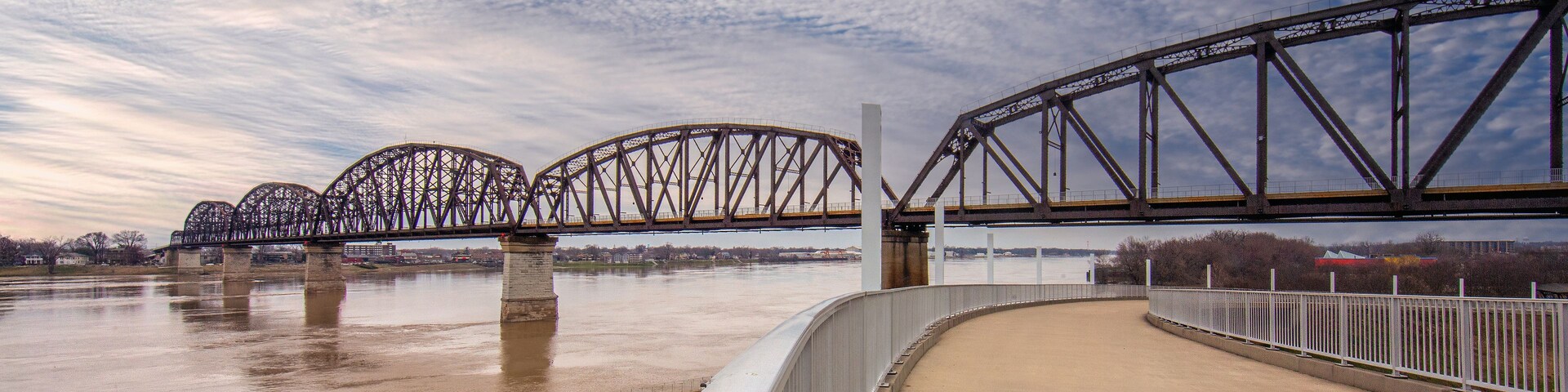 Uma ponte de aço com várias arcos sobre um rio, uma plataforma de observação de concreto e um céu nublado.