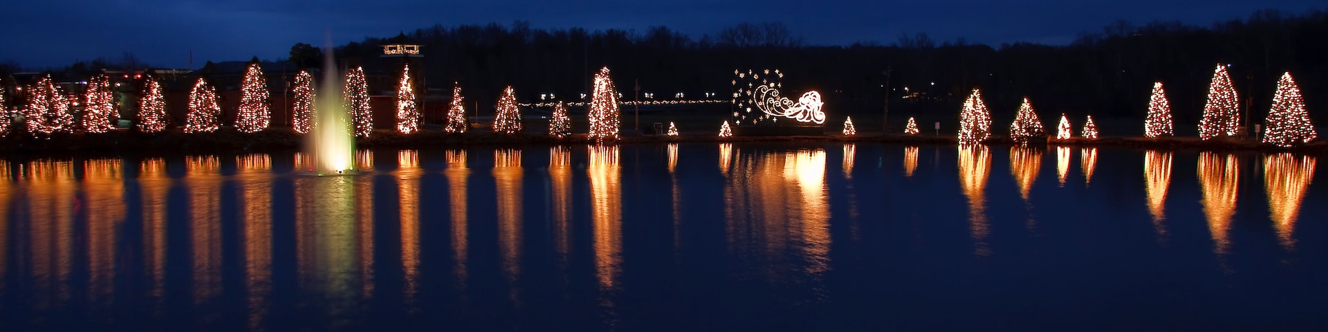 Vista noturna de um lago com árvores de Natal iluminadas na margem.