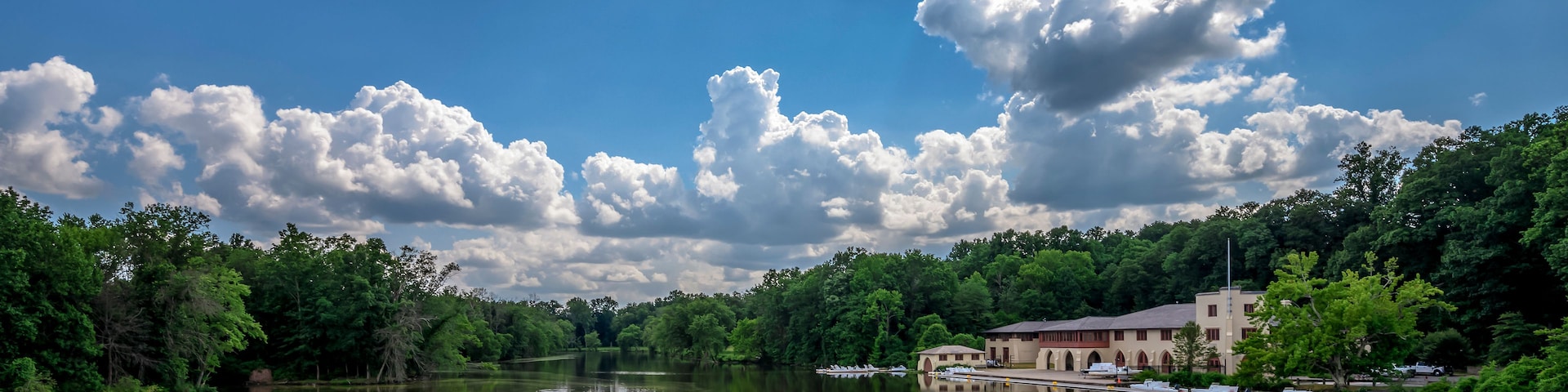 Um lago tranquilo com reflexos de nuvens, rodeado por árvores verdes densas e um edifício com telhado vermelho.