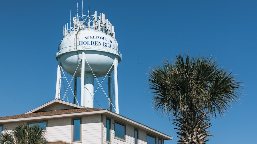 Um tanque de água com a inscrição 'WELCOME TO HOLDEN BEACH', uma palmeira e um edifício com telhado inclinado.