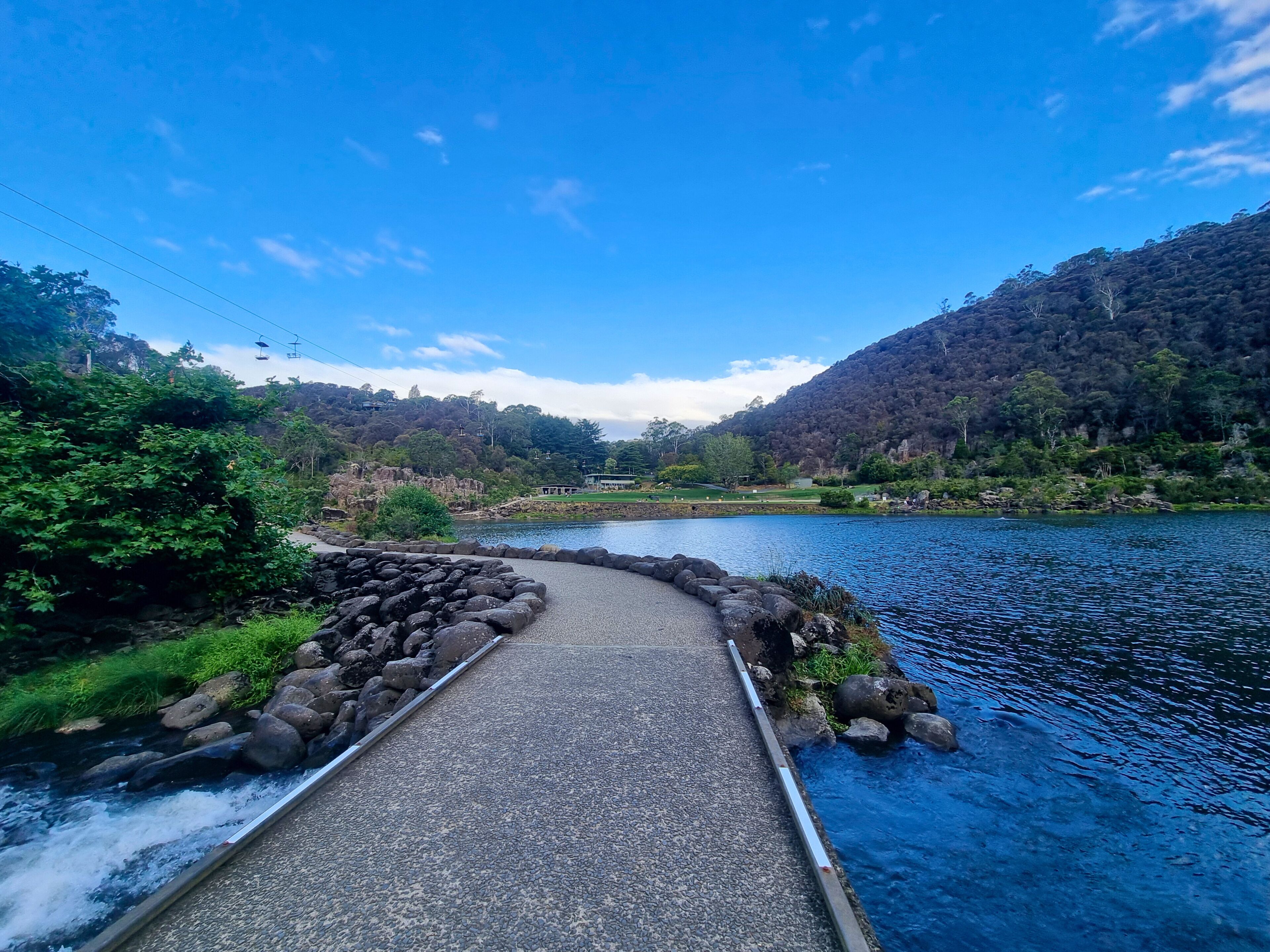 Uma trilha pavimentada ao longo de um rio, rodeada por vegetação exuberante e colinas.