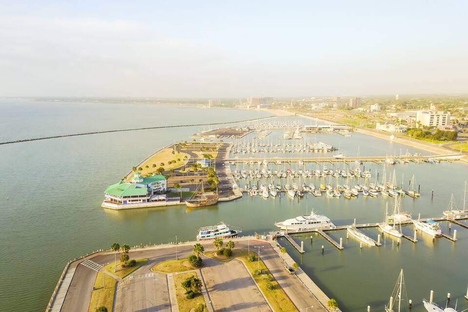 Uma marina com barcos atracados, um grande navio de cruzeiro e uma estrada que leva a um edifício com telhado verde.