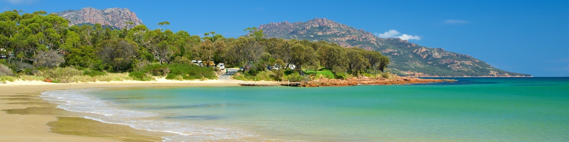 Uma praia com águas turquesa translúcidas, areal arenoso e um cenário de árvores verdes e montanhas rochosas.