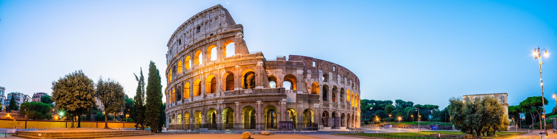O Coliseu em Roma, Itália, ao anoitecer, com árvores ao redor e uma praça pavimentada.