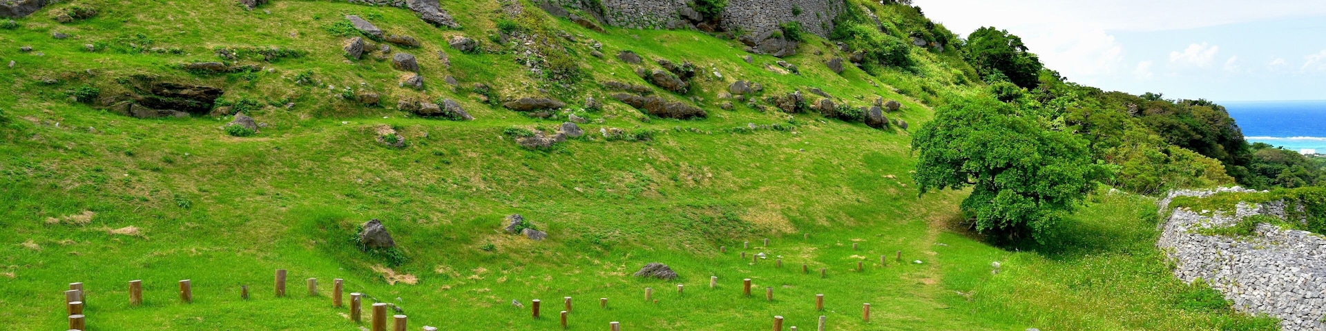 Uma colina gramada com ruínas de pedra e estacas de madeira, provavelmente um local histórico.