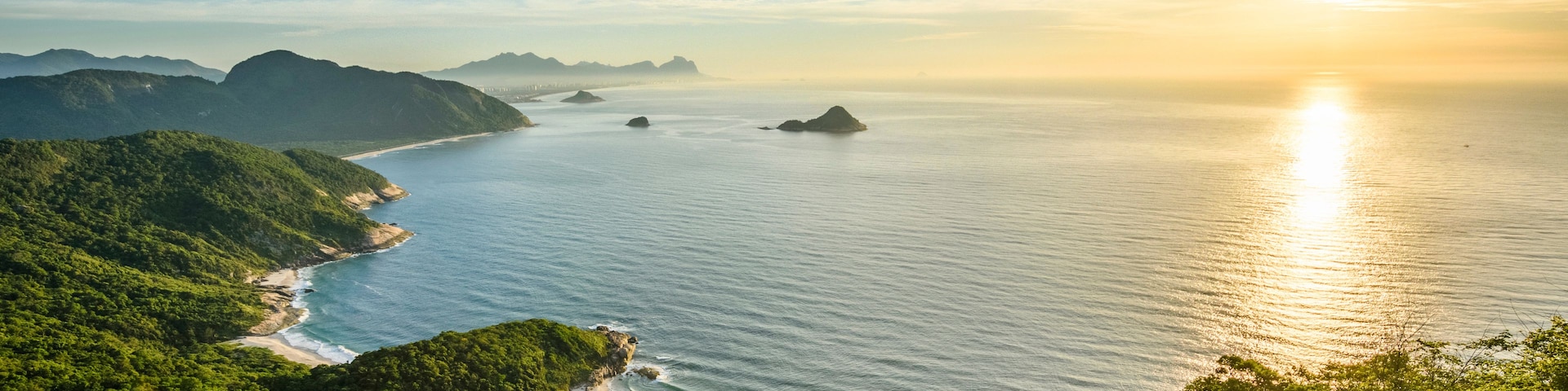 Paisagem costeira com céu limpo, sol se pondo ou nascendo sobre o oceano, e terreno montanhoso com vegetação densa.