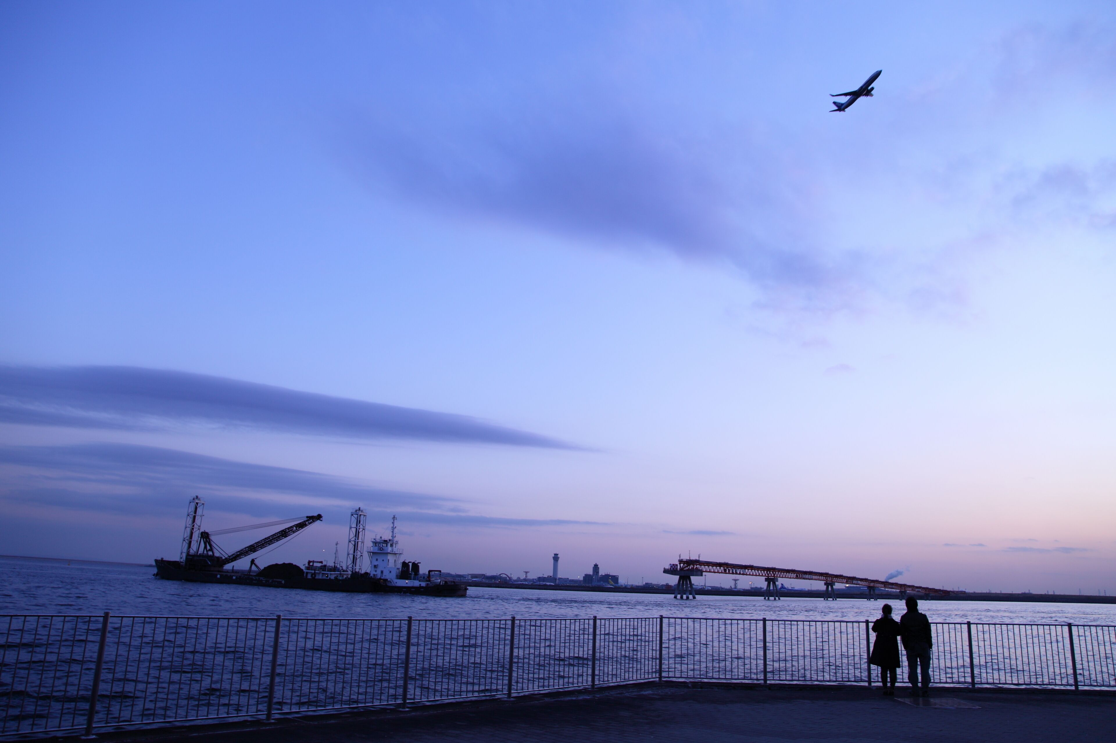 Um pôr do sol sobre um rio com uma ponte, um barco e uma pessoa perto do corrimão.