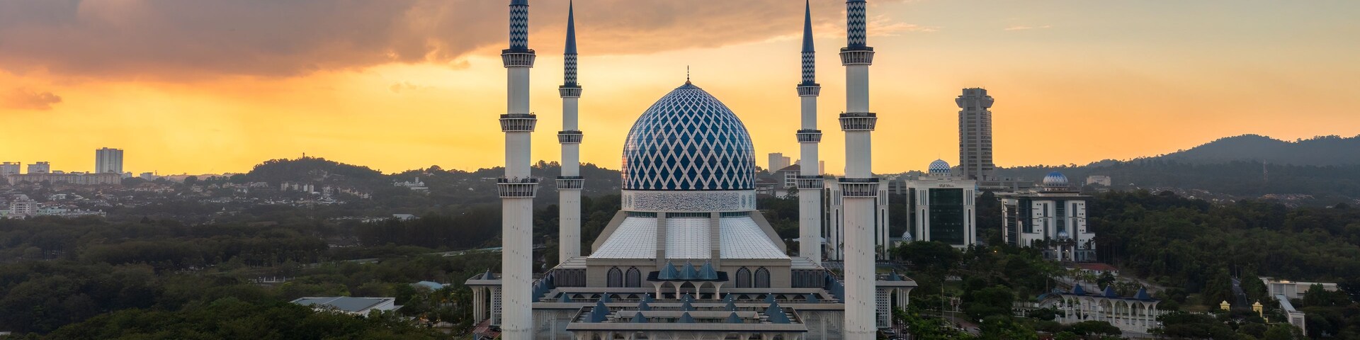 Uma mesquita com uma grande cúpula central e quatro minaretes, rodeada por árvores e edifícios urbanos.