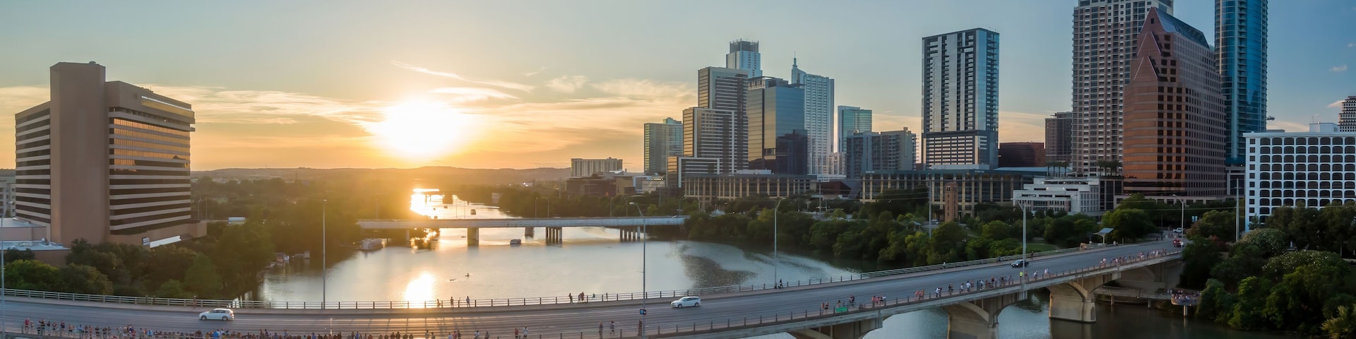 Silhueta urbana ao pôr do sol, com um rio, uma ponte e edifícios modernos.