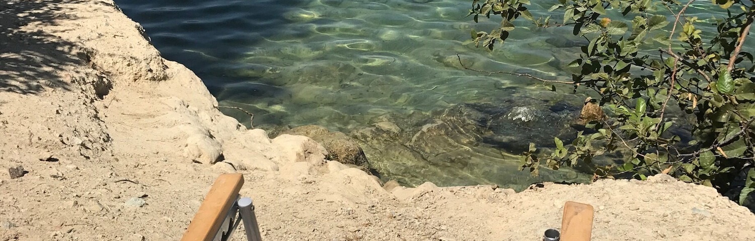 Uma cadeira de praia com revistas e um bastão de madeira numa margem arenosa à beira de um lago calmo, com montanhas ao fundo.