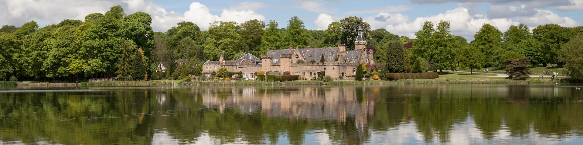 Um edifício grande e histórico com uma torre, rodeado por vegetação exuberante e um lago tranquilo que reflete a estrutura.