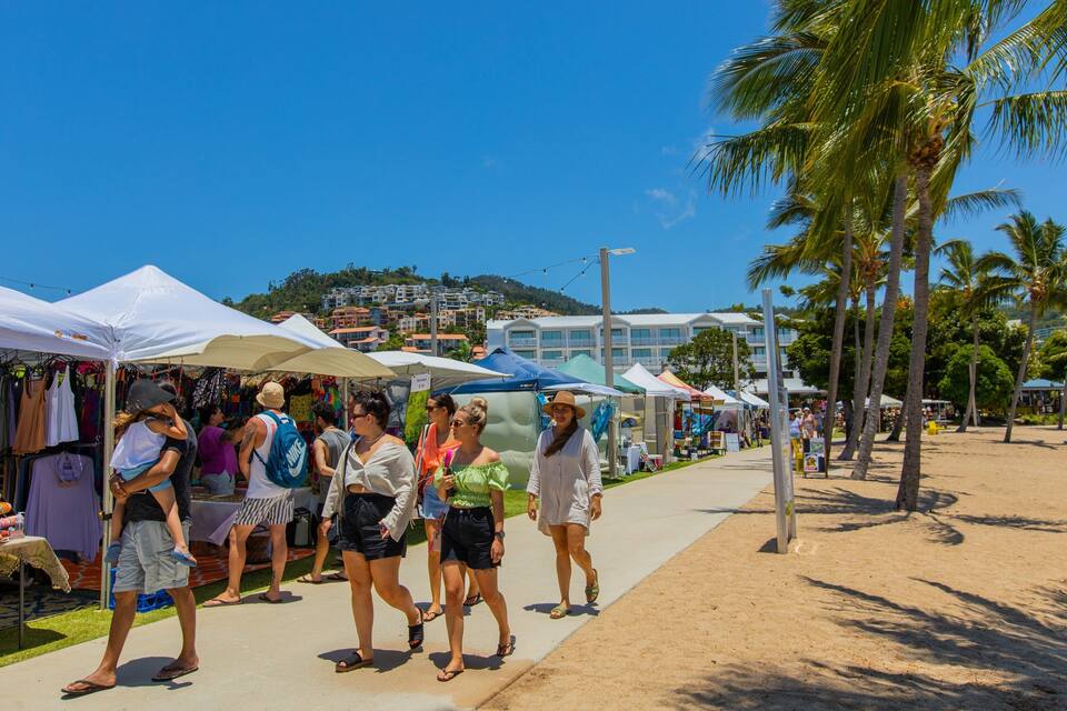 Um mercado ensolarado à beira-mar, com pessoas caminhando e barracas vendendo roupas.
