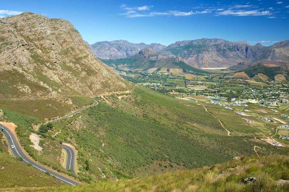 Uma estrada sinuosa serpenteia pela montanha, um vale com casas dispersas e uma cadeia de montanhas ao fundo.