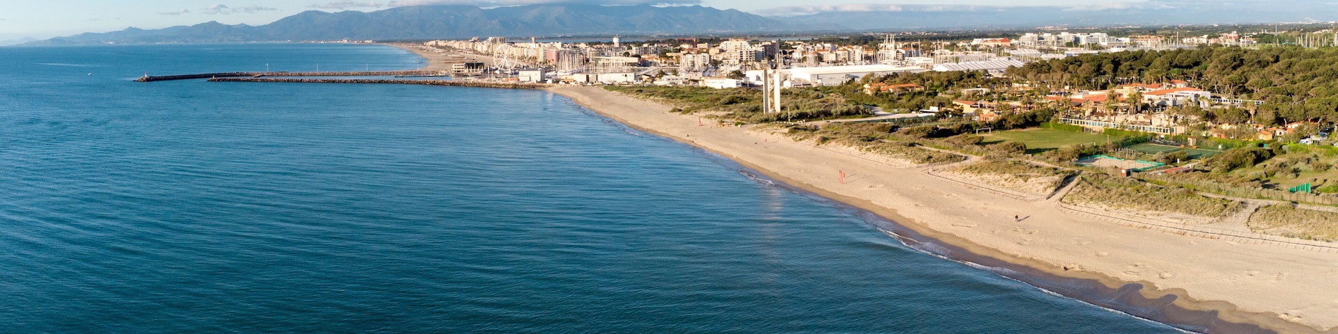 Uma cidade costeira com uma extensa praia, um cais que se projeta para o mar e montanhas ao longe.