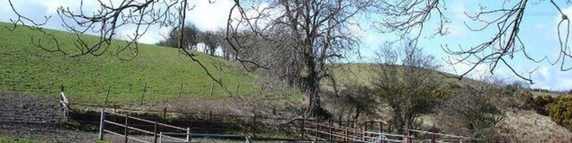 Paisagem rural com um pequeno riacho, cerca de madeira e uma colina ao fundo.