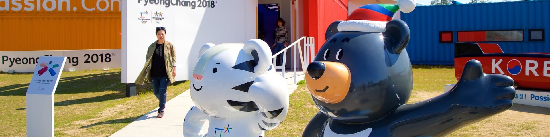 Dois mascotes, um urso branco e um urso azul, em frente a um edifício com uma placa que diz 'PyeongChang 2018'.