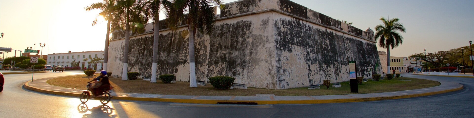 Uma fortaleza histórica com uma torre de sino, palmeiras e uma rotatória com placa de limite de velocidade.
