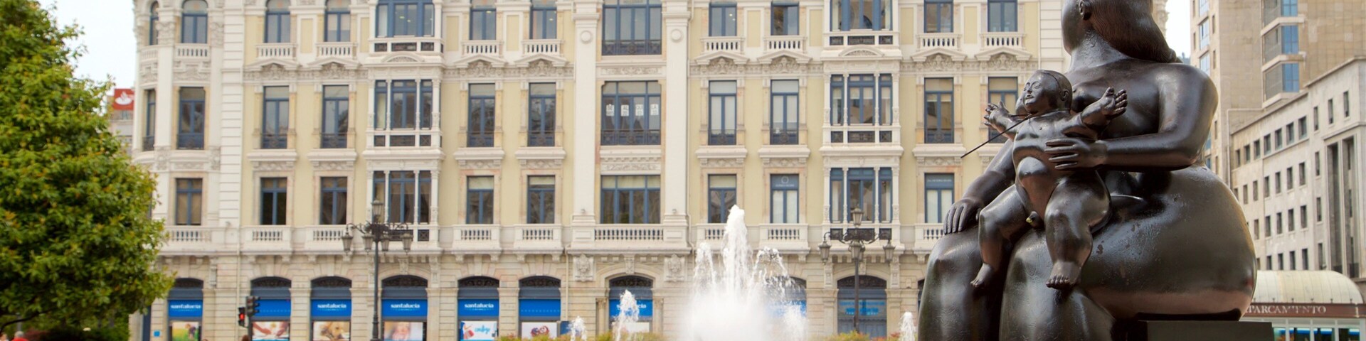 Estátua de uma mulher segurando uma criança em frente a um edifício grande e ricamente decorado, com uma fonte no centro.