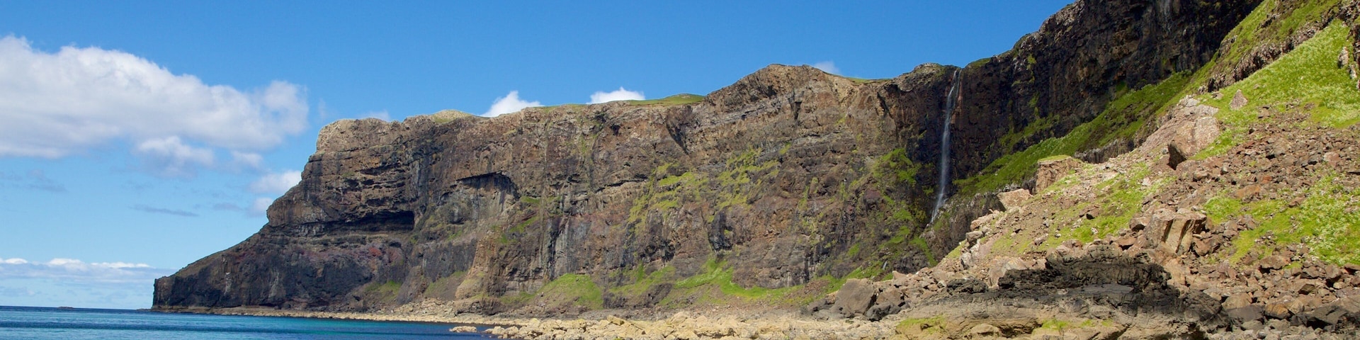 Paisagem costeira com um penhasco rochoso, uma cascata e um mar azul e límpido.