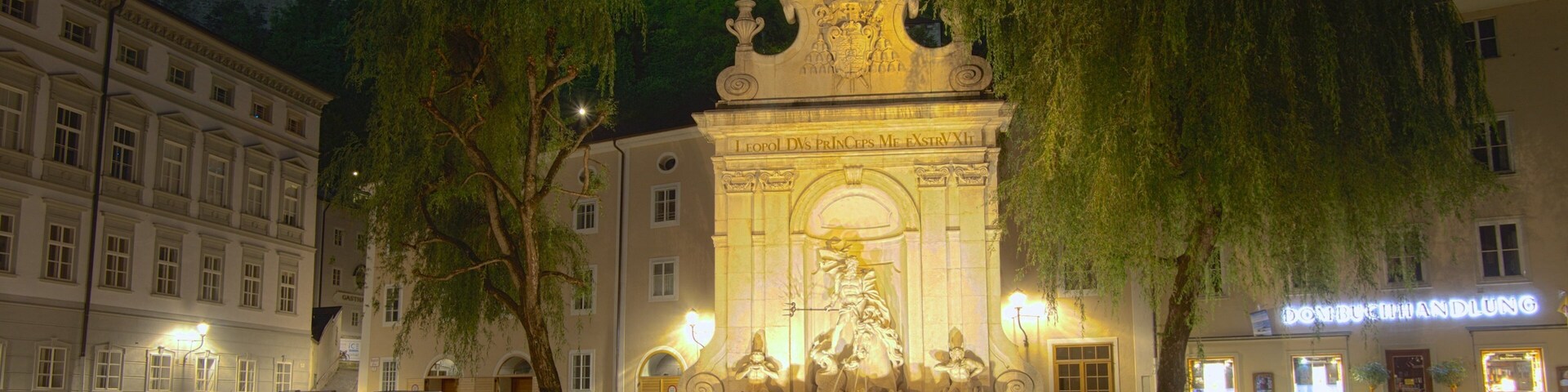 Vista noturna de uma fonte histórica com estátua, cercada por árvores e um castelo ao fundo.