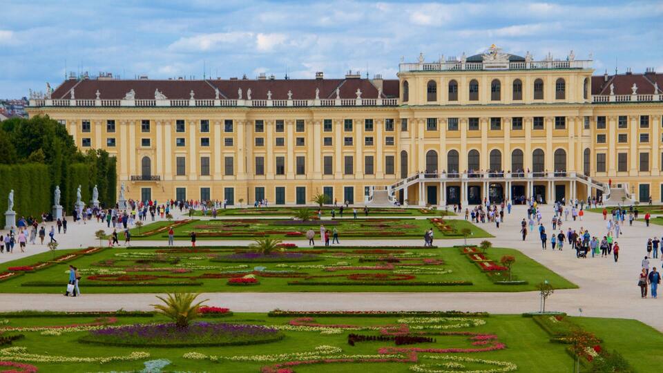 Um grande edifício clássico com fachada simétrica, uma escadaria central e um jardim formal com canteiros de flores geométricos.