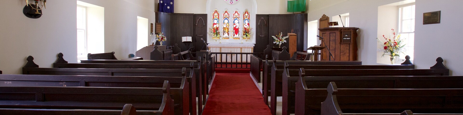 Interior de igreja com bancos de madeira, um corredor com tapete vermelho, vitrais e bandeiras.