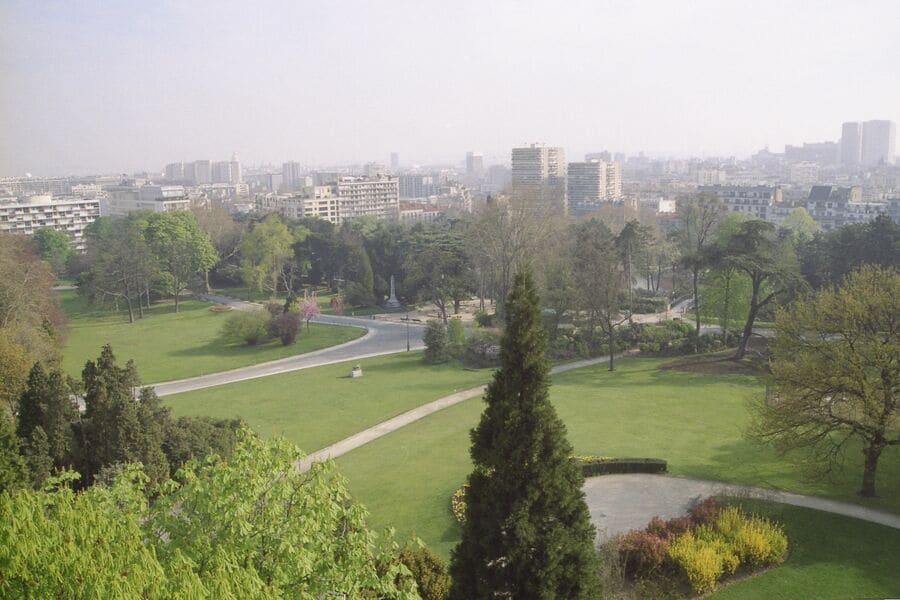 Um parque com um caminho serpenteante, rodeado por árvores e edifícios ao fundo.