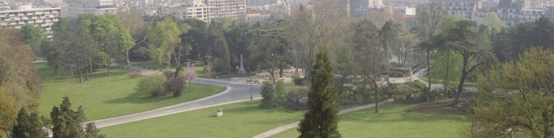 Um parque com um caminho serpenteante, rodeado por árvores e edifícios ao fundo.