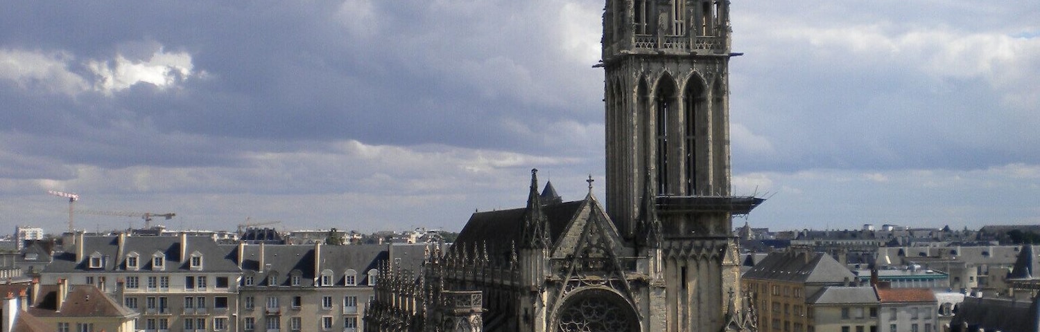 Uma catedral histórica com uma torre alta, rodeada por edifícios urbanos e uma rua movimentada.