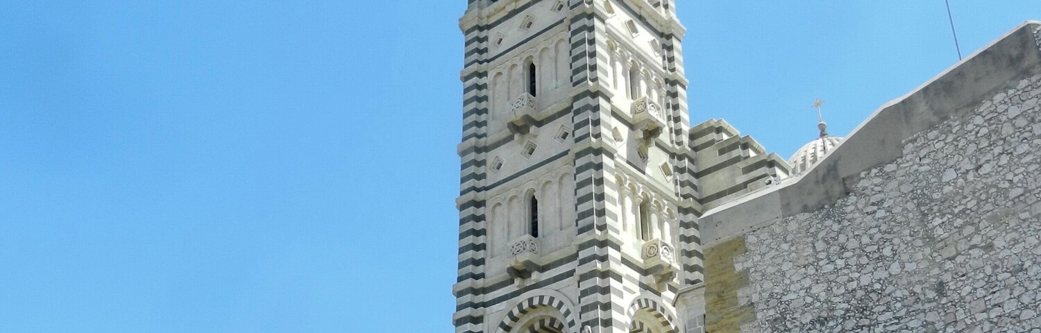 Uma torre histórica com trabalho em pedra intrincado e estátuas, uma escadaria de pedra que leva até ela, e um céu azul claro ao fundo.